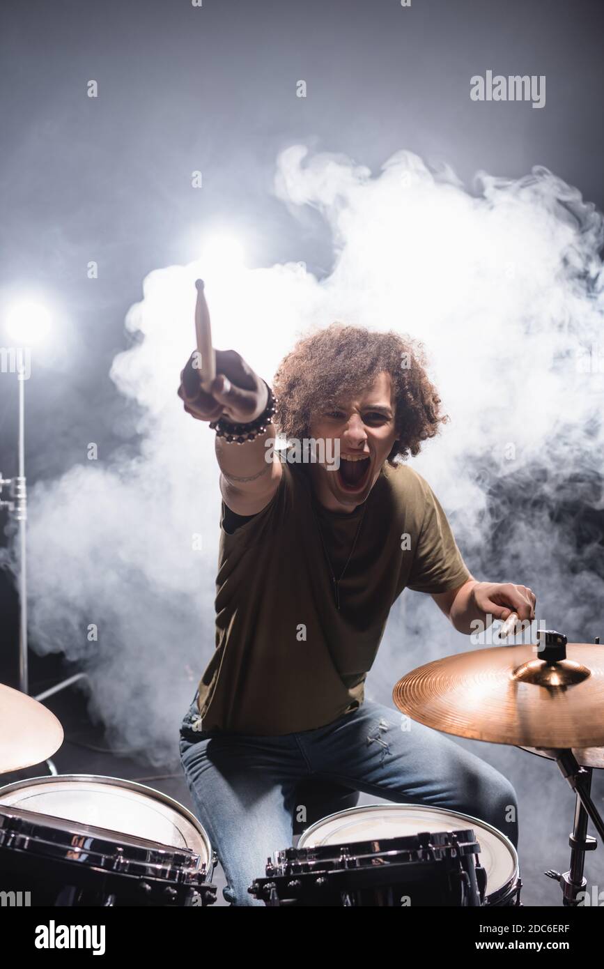 Curly drummer pointing with drumstick while shouting and sitting at ...