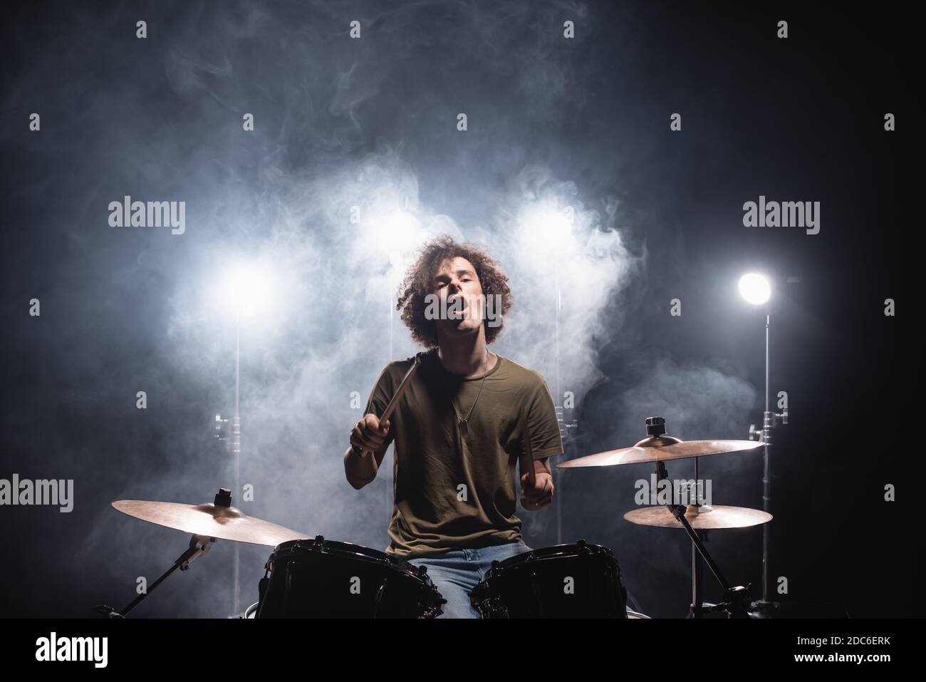 Excited curly drummer with open mouth sitting at drum kit with smoke ...