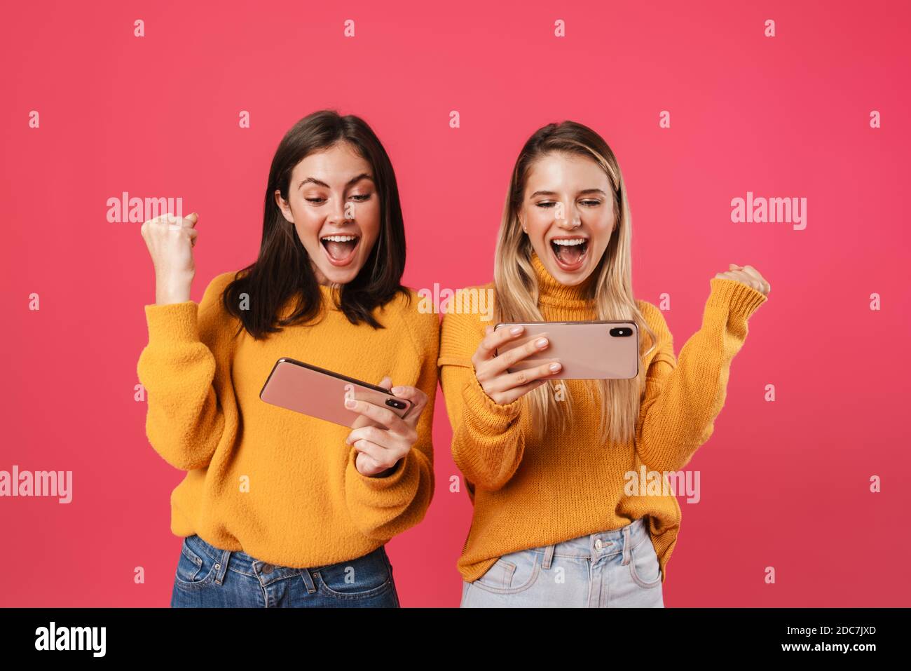 Two happy pretty girls celebrating success while using mobile phones ...