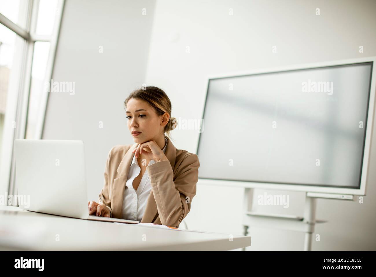 Pretty young woman working on laptop in bright office with big screen ...