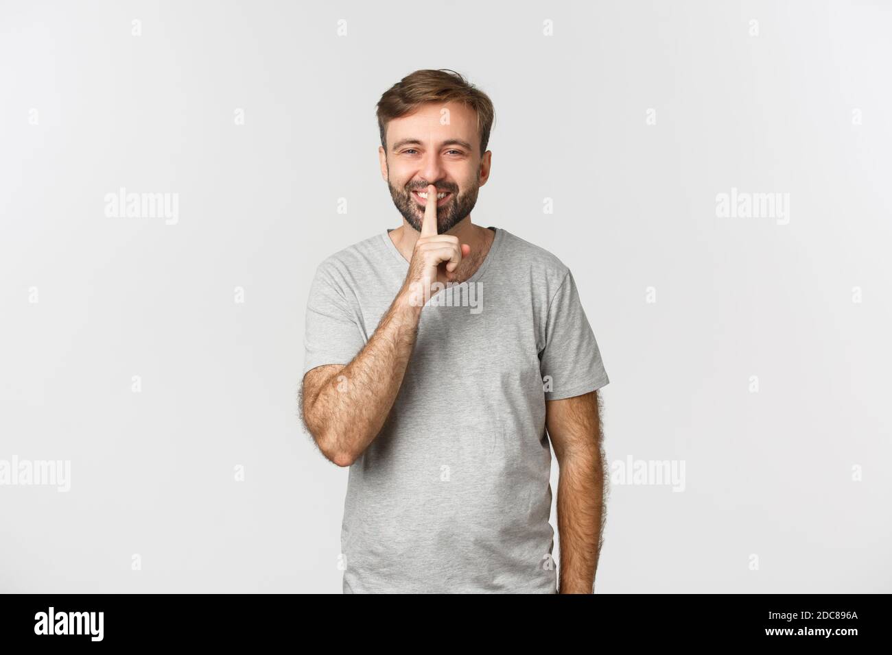 Portrait of handsome smiling man in grey t-shirt, hushing at you kindly ...
