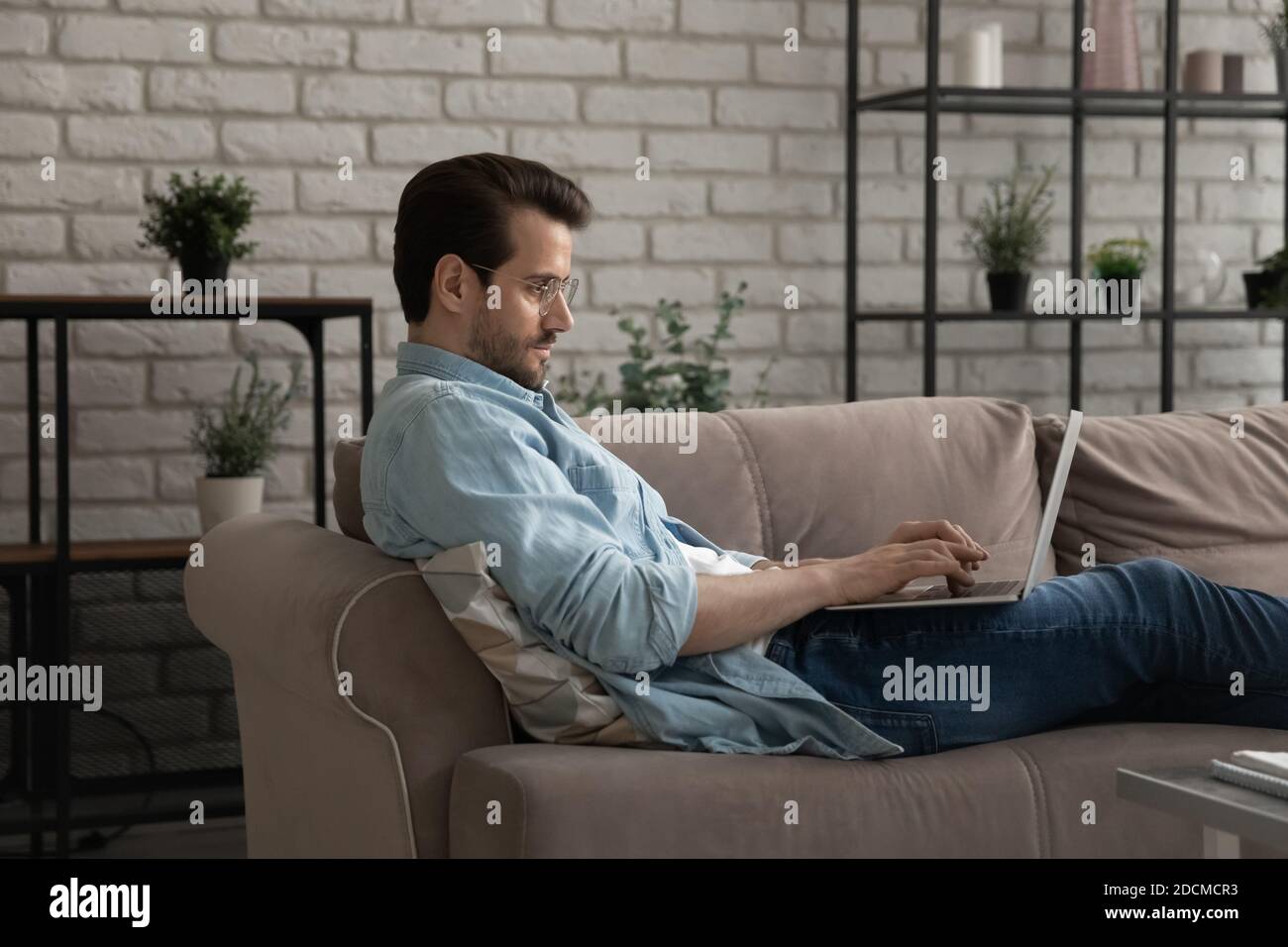 Relaxed young man lying on sofa with computer on laps Stock Photo - Alamy
