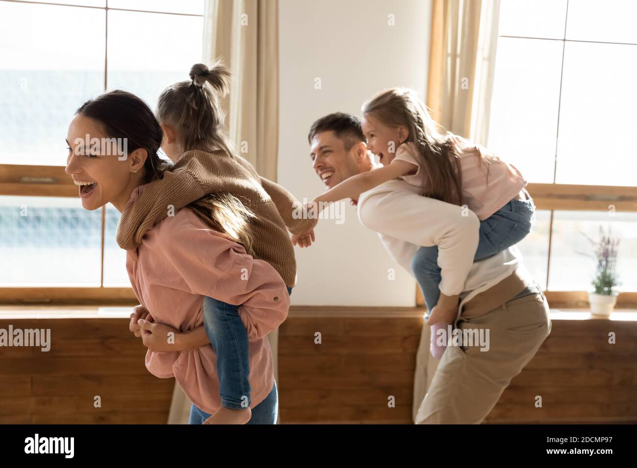 Cheerful couple piggyback riding small daughters play together at home ...
