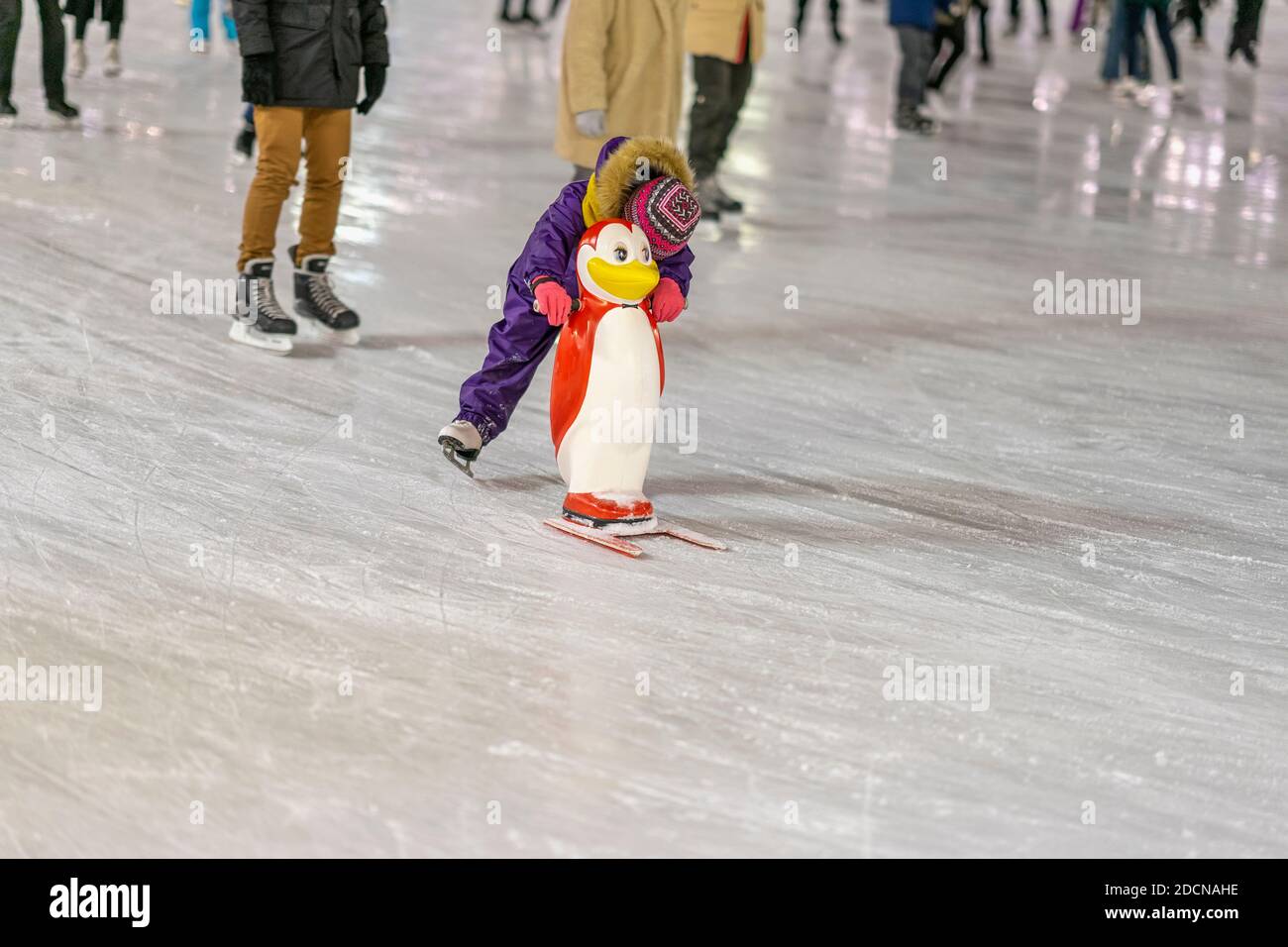 Ice rink. Little child holds a penguin for support and learn to skate ...