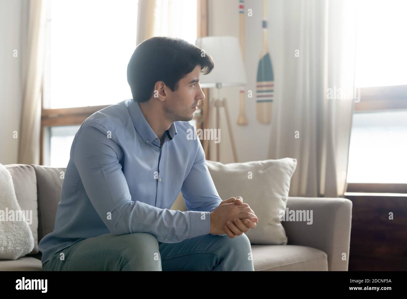 Thoughtful male sitting on couch alone looking at distance thinking ...