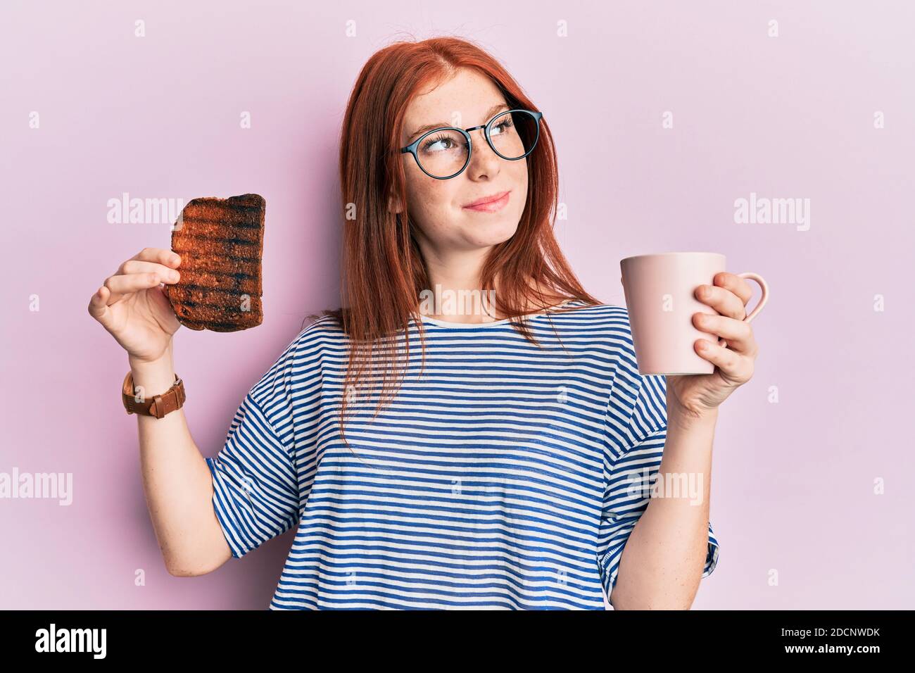 Young red head girl holding burned toast for breakfast smiling looking ...