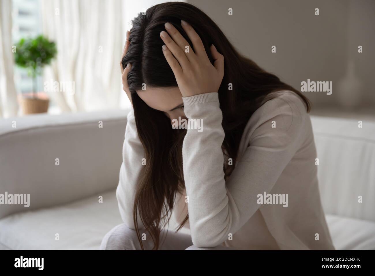 Stressed confused young female sitting on sofa suffering hugging head ...