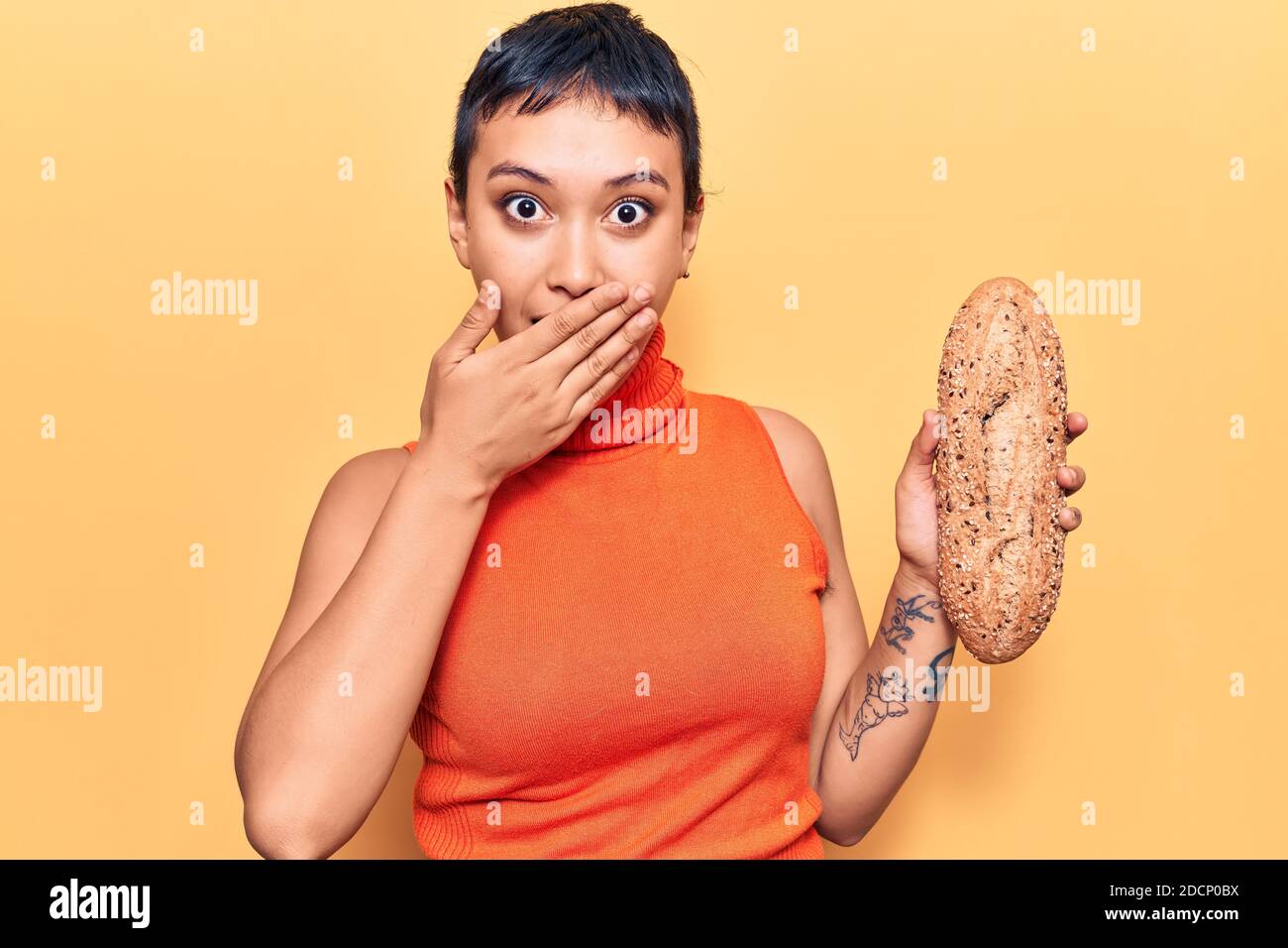 Young woman holding wholemeal bread covering mouth with hand, shocked ...