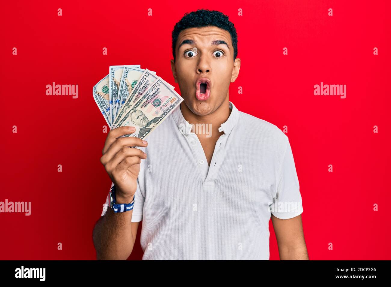 Young arab man holding dollars scared and amazed with open mouth for ...