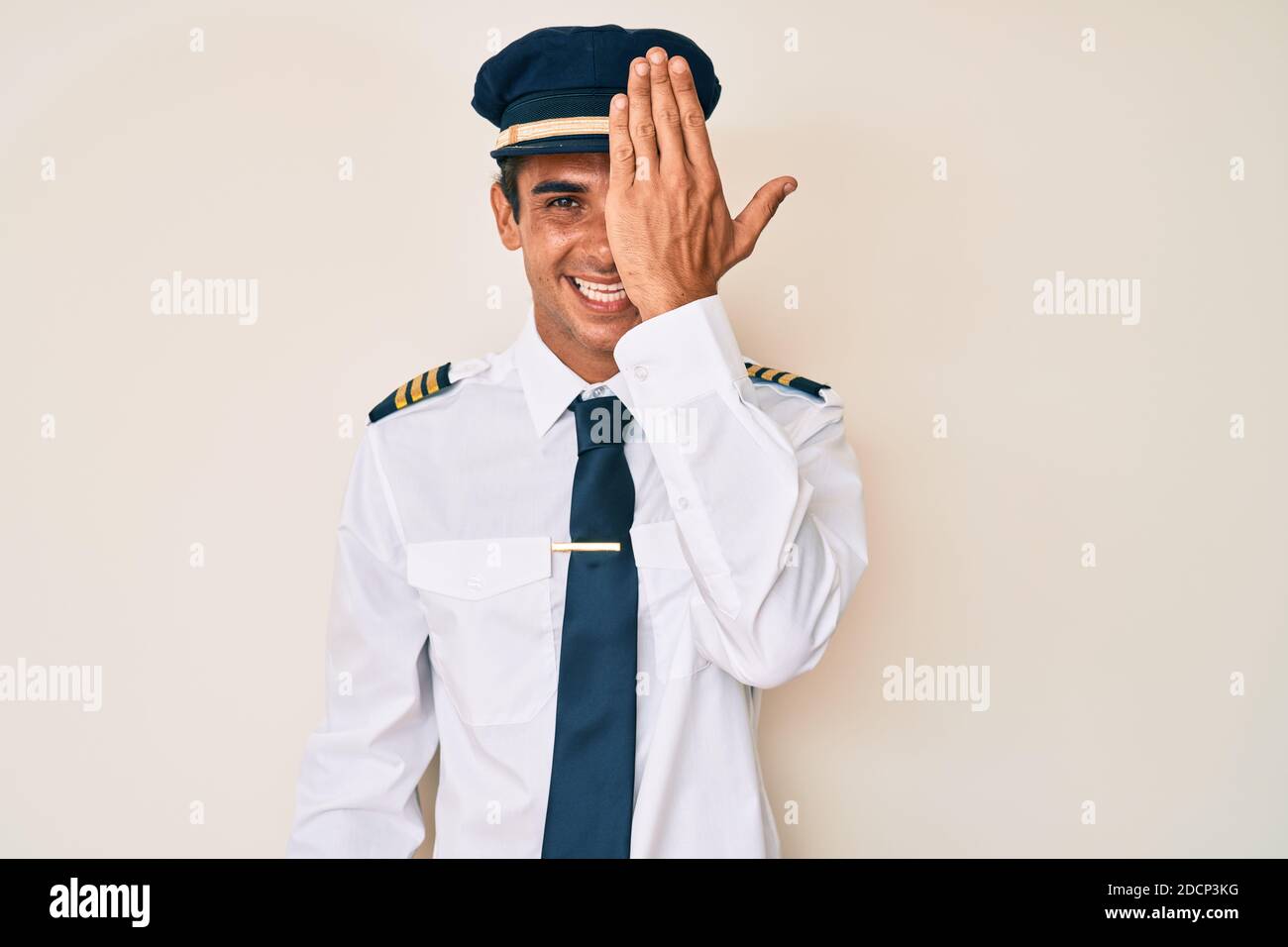 Young hispanic man wearing airplane pilot uniform covering one eye with ...