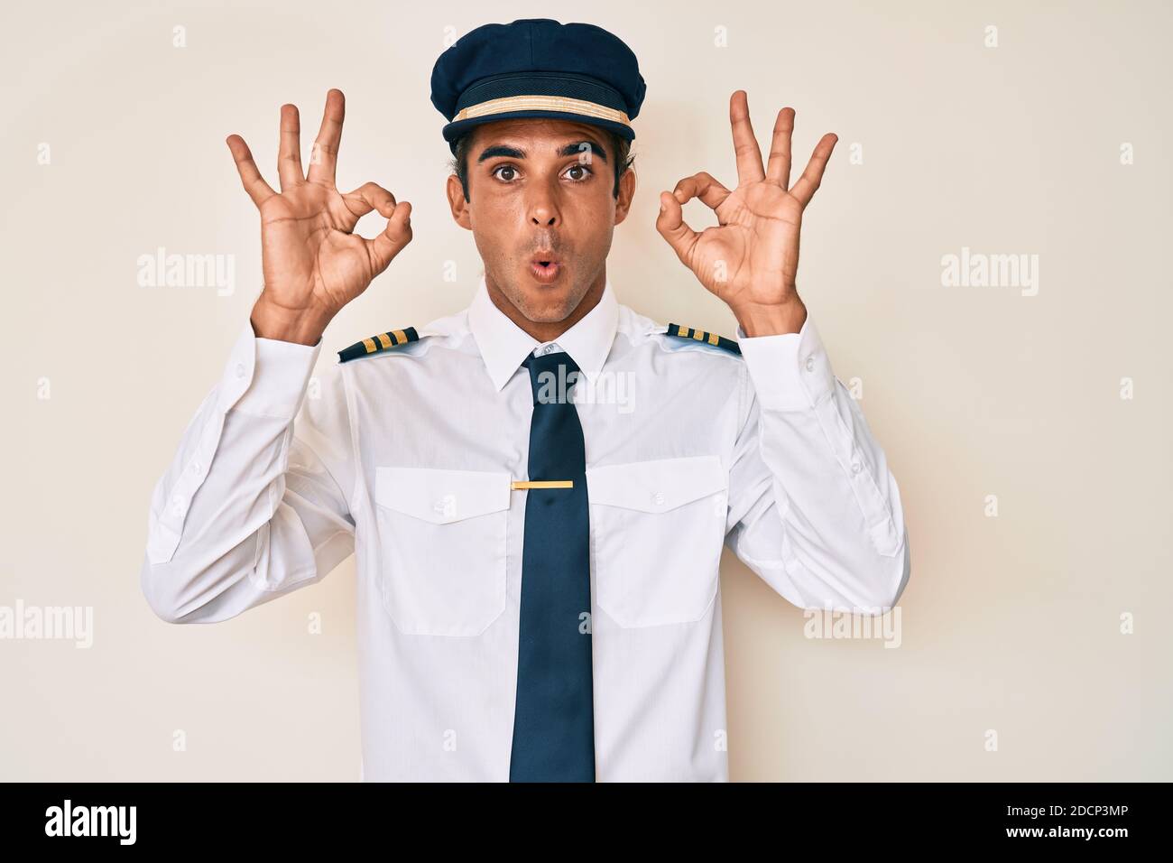 Young hispanic man wearing airplane pilot uniform looking surprised and ...