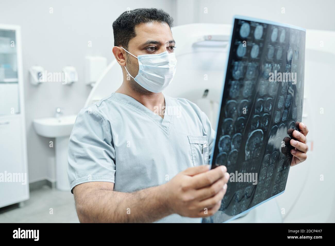 Serious male radiologist in mask and uniform looking at x-ray image of ...