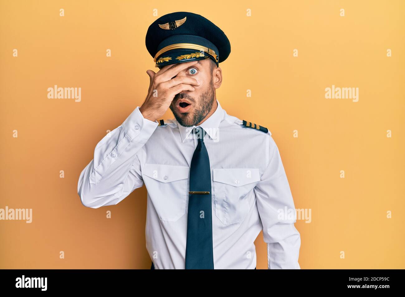 Handsome hispanic man wearing airplane pilot uniform peeking in shock ...