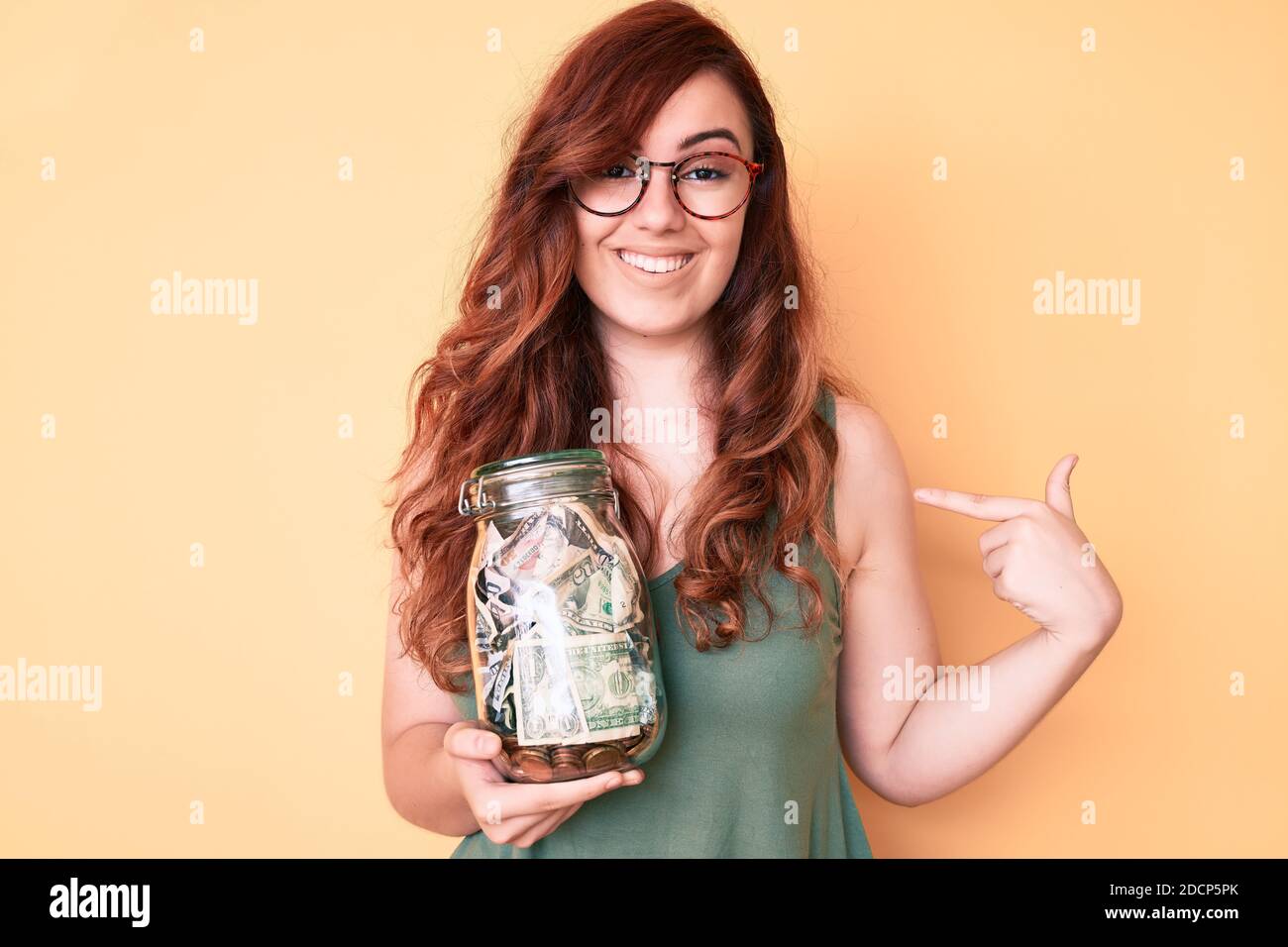 Young beautiful woman wearing glasses holding jar with savings pointing ...