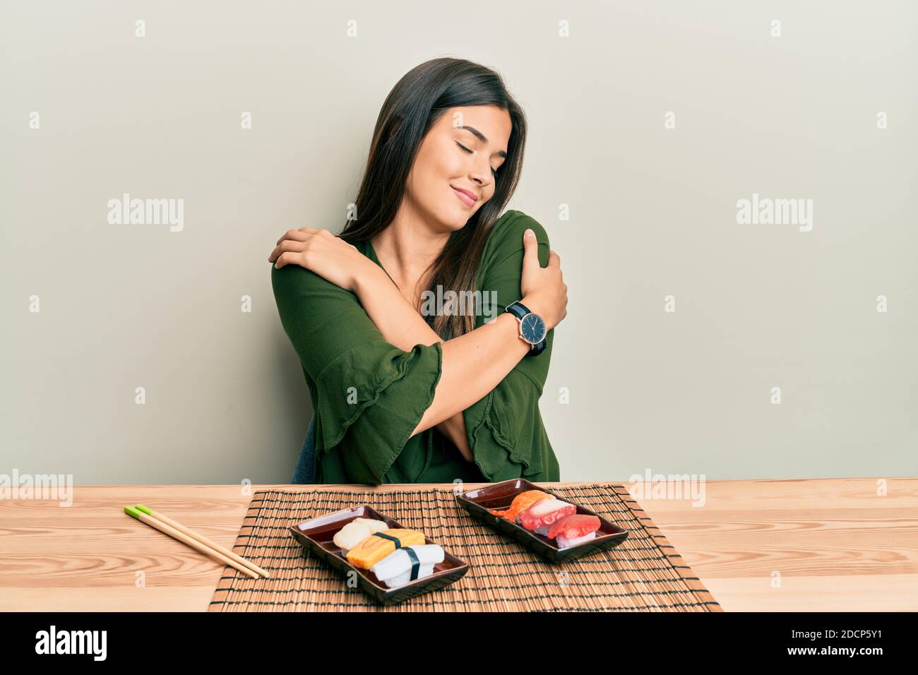 Young brunette woman eating sushi sitting on the table hugging oneself ...