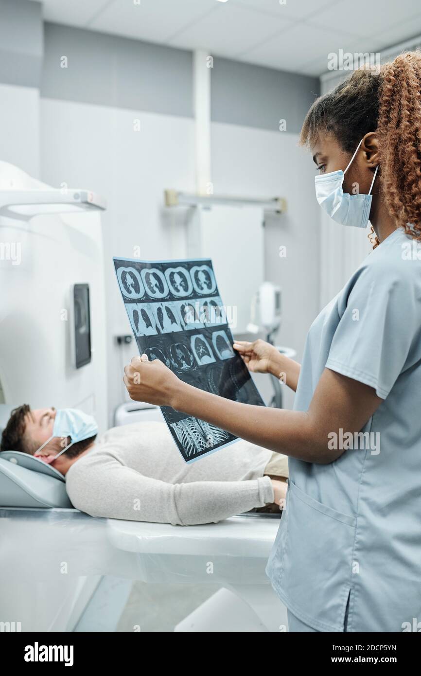 Young African female radiologist in uniform and mask analyzing x-ray ...