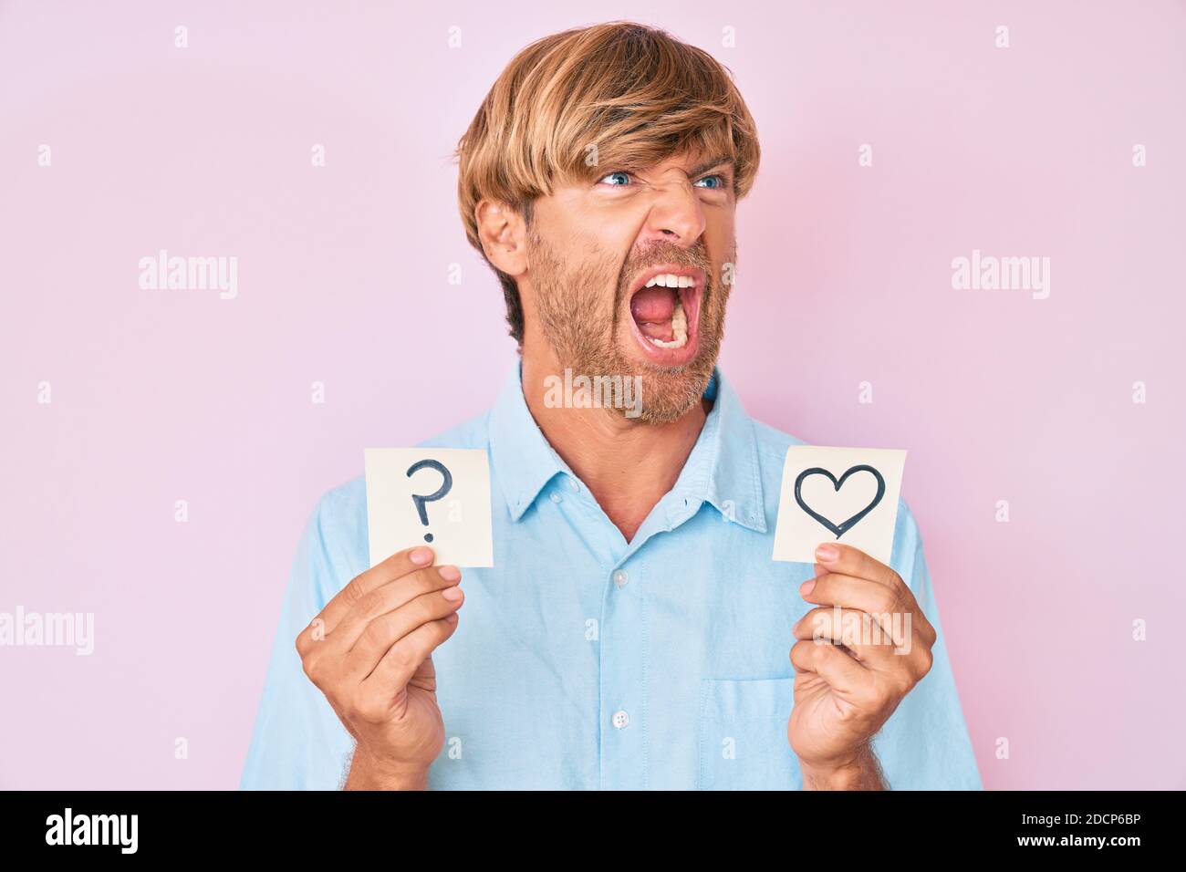 Young blond man holding heart and question mark reminder angry and mad ...
