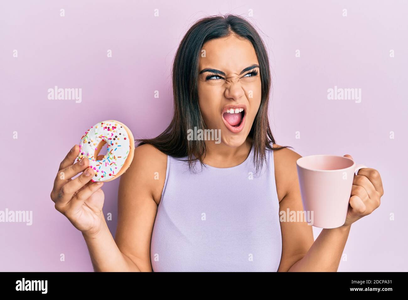 Young brunette woman eating doughnut and drinking coffee angry and mad ...