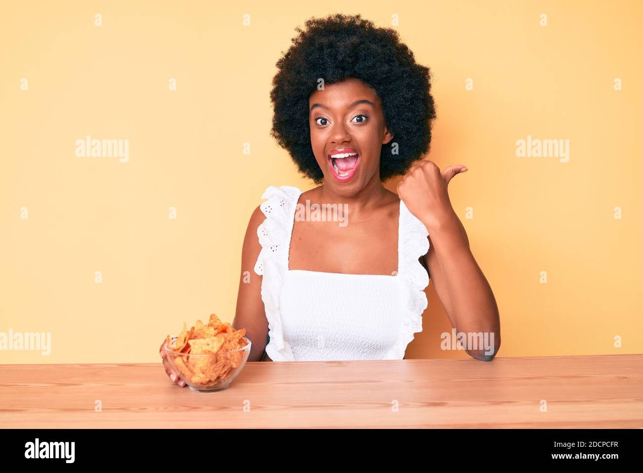 Young african american woman holding nachos potato chips pointing thumb ...