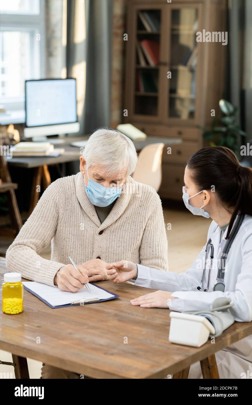 Sick senior male patient in mask and casualwear signing medical paper ...