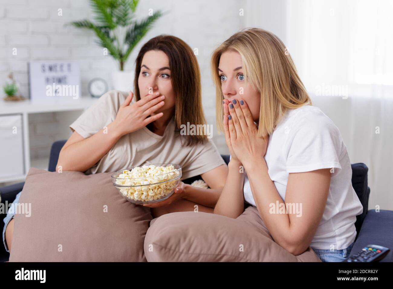 two surprised young women eating popcorn and watching something on tv ...