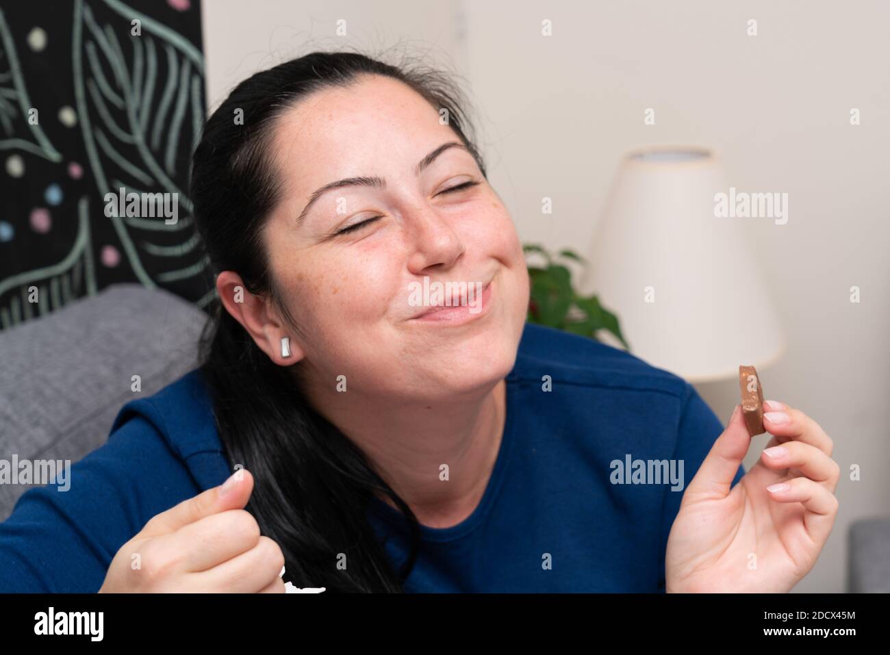 Cheerful adult female model eating milk chocolate as guilty pleasure ...