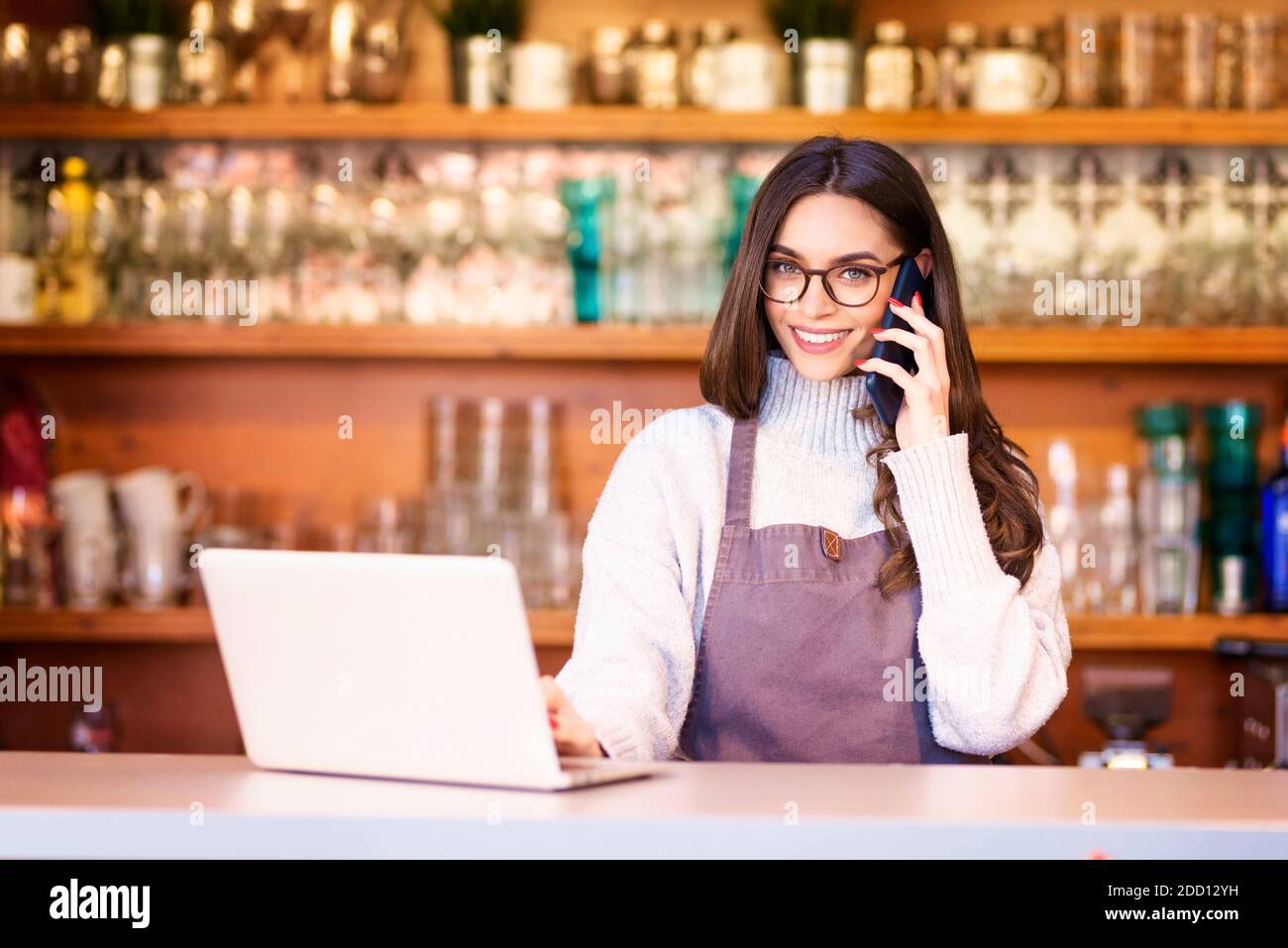 Portrait of a beautiful waitress using mobile phone and laptop while ...
