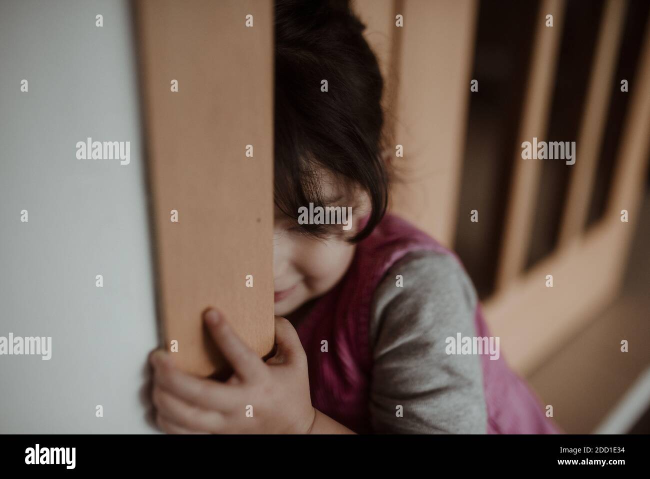 A closeup of a cute Caucasian girl hiding next to the wooden door frame ...