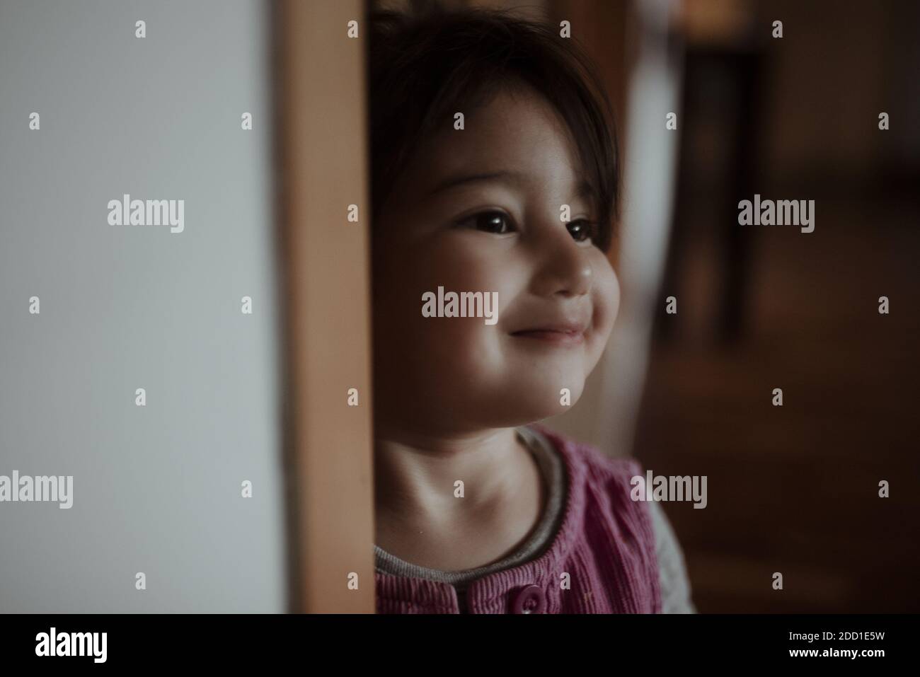 A closeup of a cute Caucasian girl hiding next to the wooden door frame ...