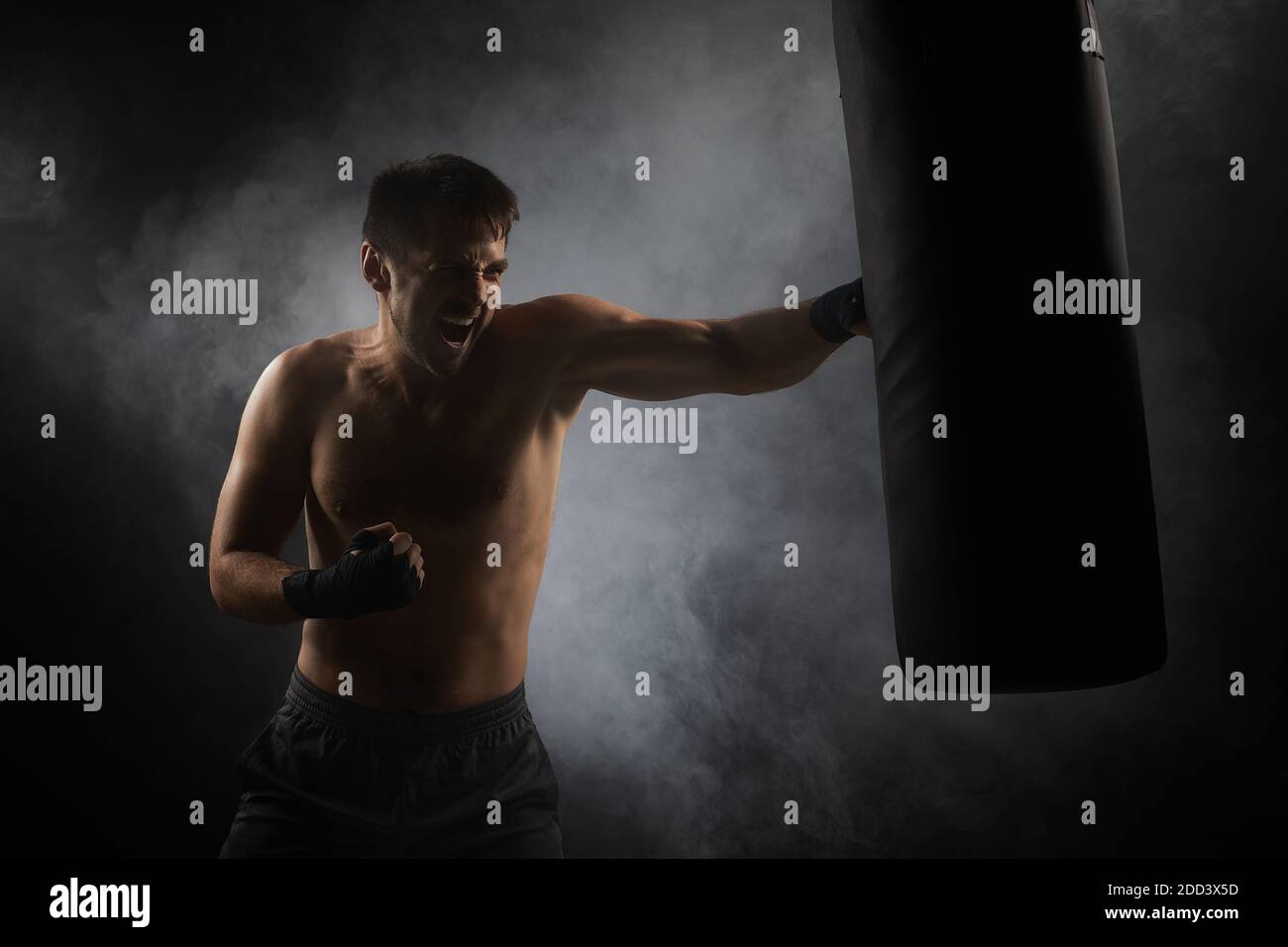 Aggressive boxer in black boxing wraps punching in boxing bag on dark ...