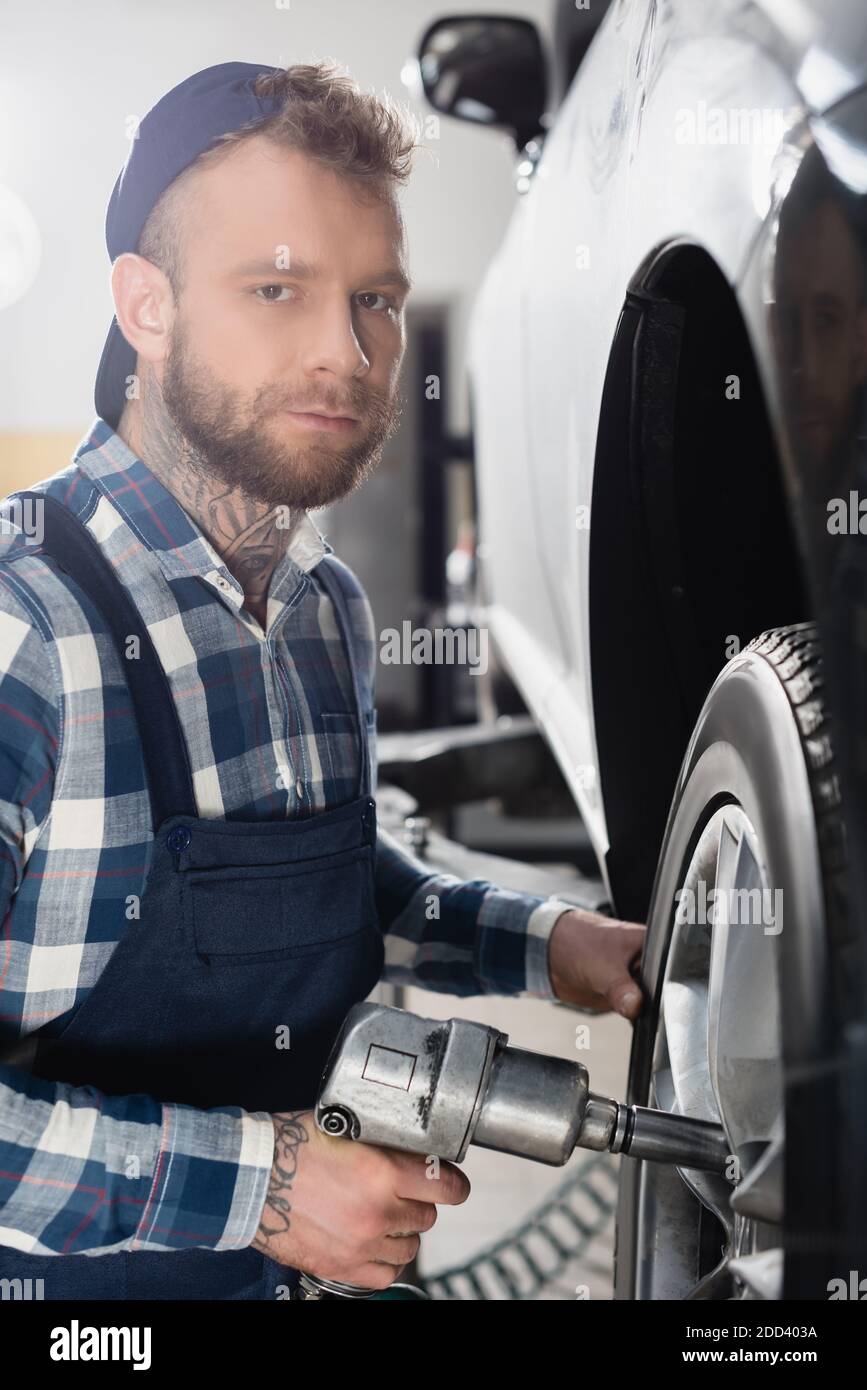 young mechanic looking at camera while fixing car wheel with pneumatic ...