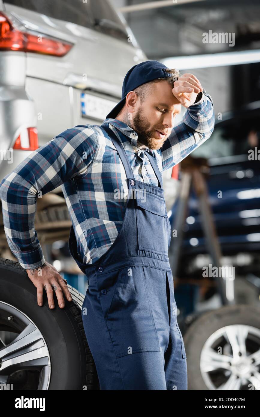 exhausted mechanic holding hand near forehead while standing near car ...