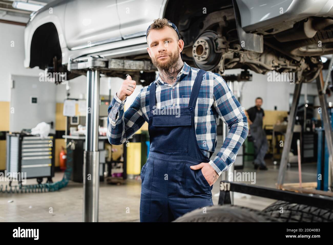 young mechanic showing thumb up while holding hand in pocket near ...