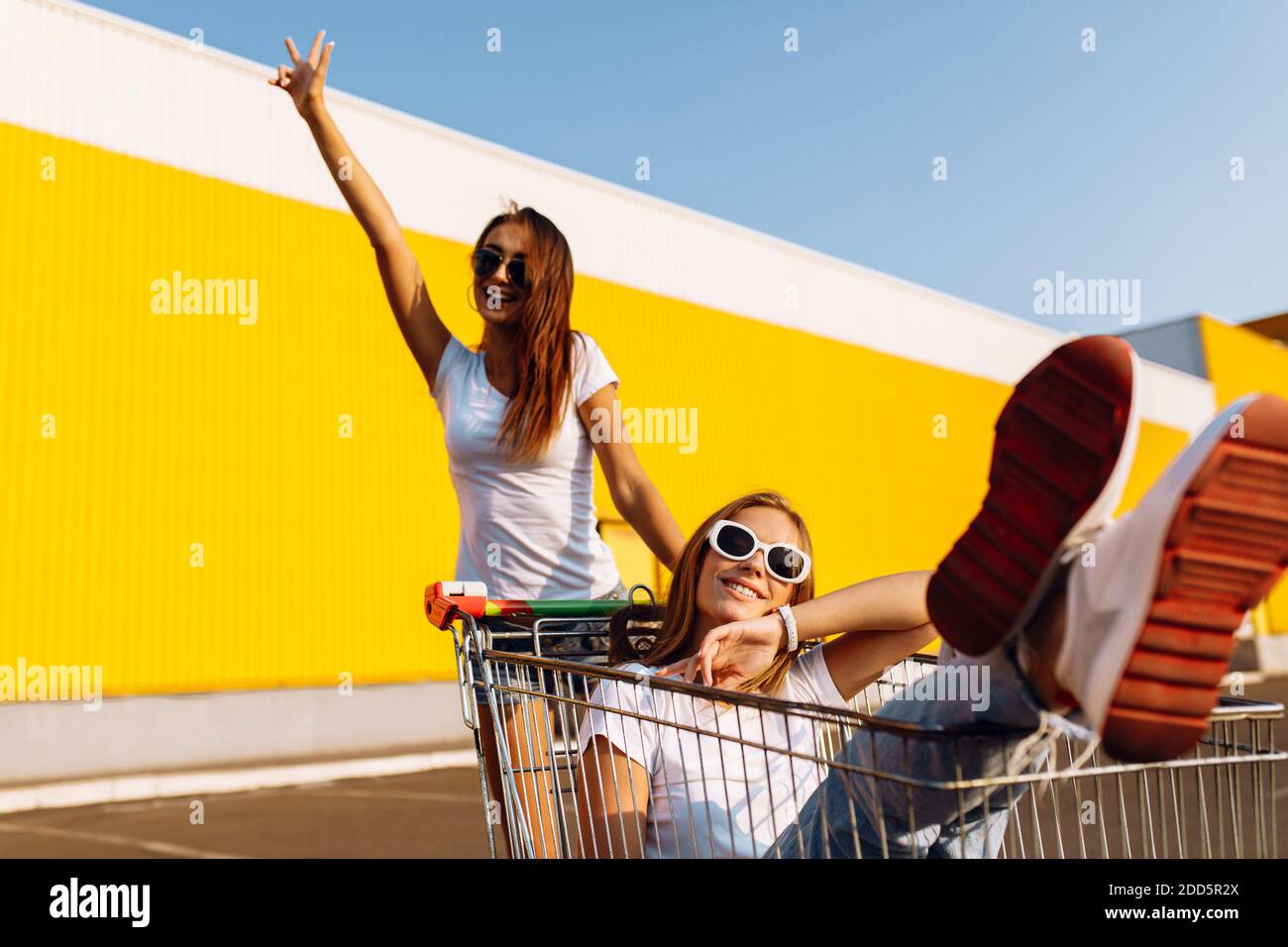 Excited young women ride shopping cart in city against yellow wall of ...