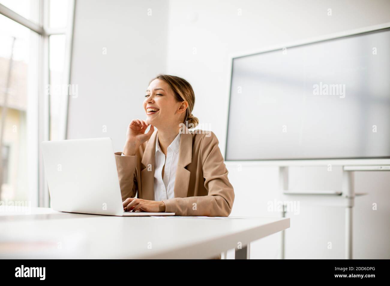 Pretty young woman working on laptop in bright office with big screen ...