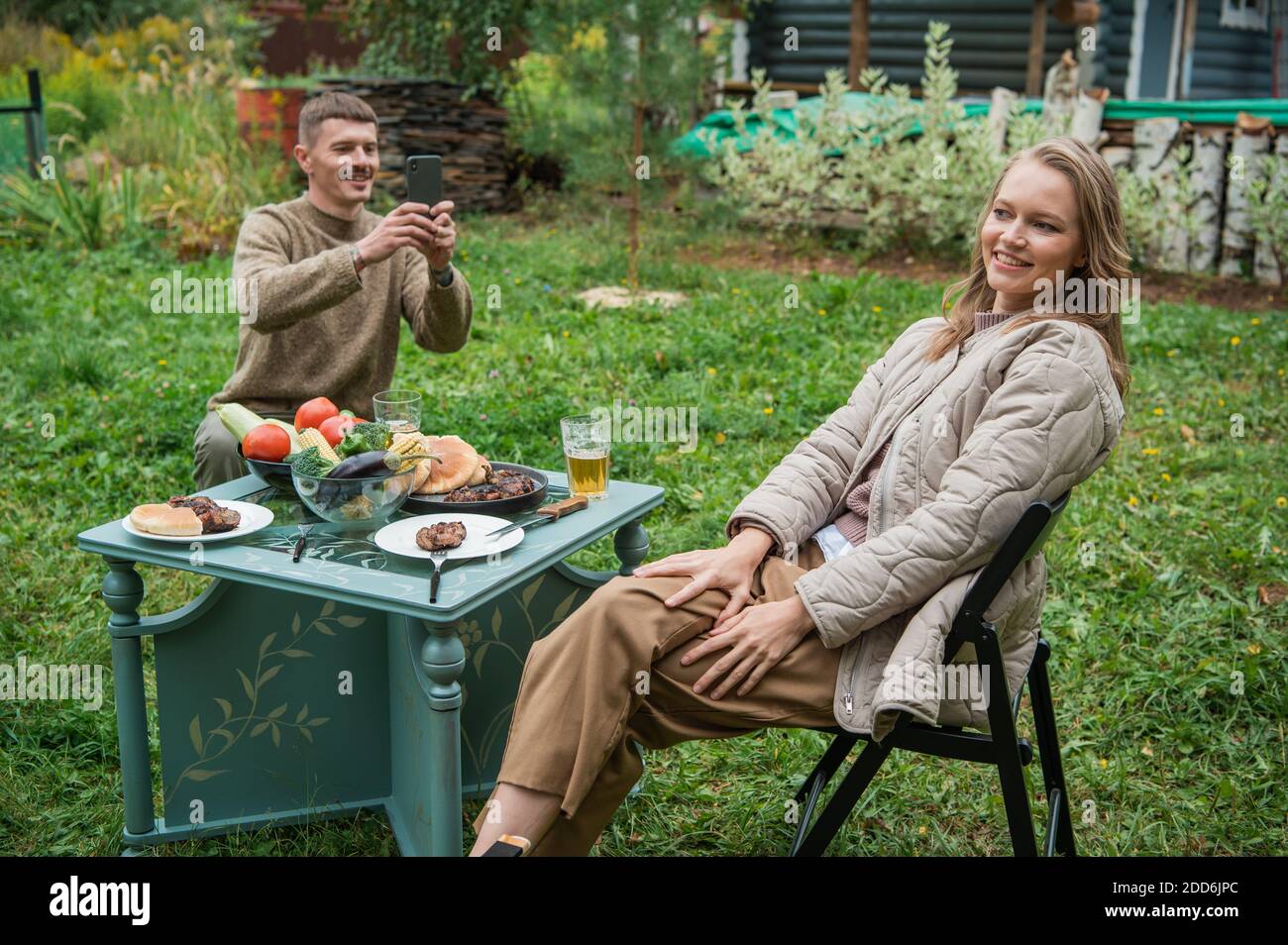 A young man takes photos of his girlfriend on a smartphone during a picnic in nature near their ...