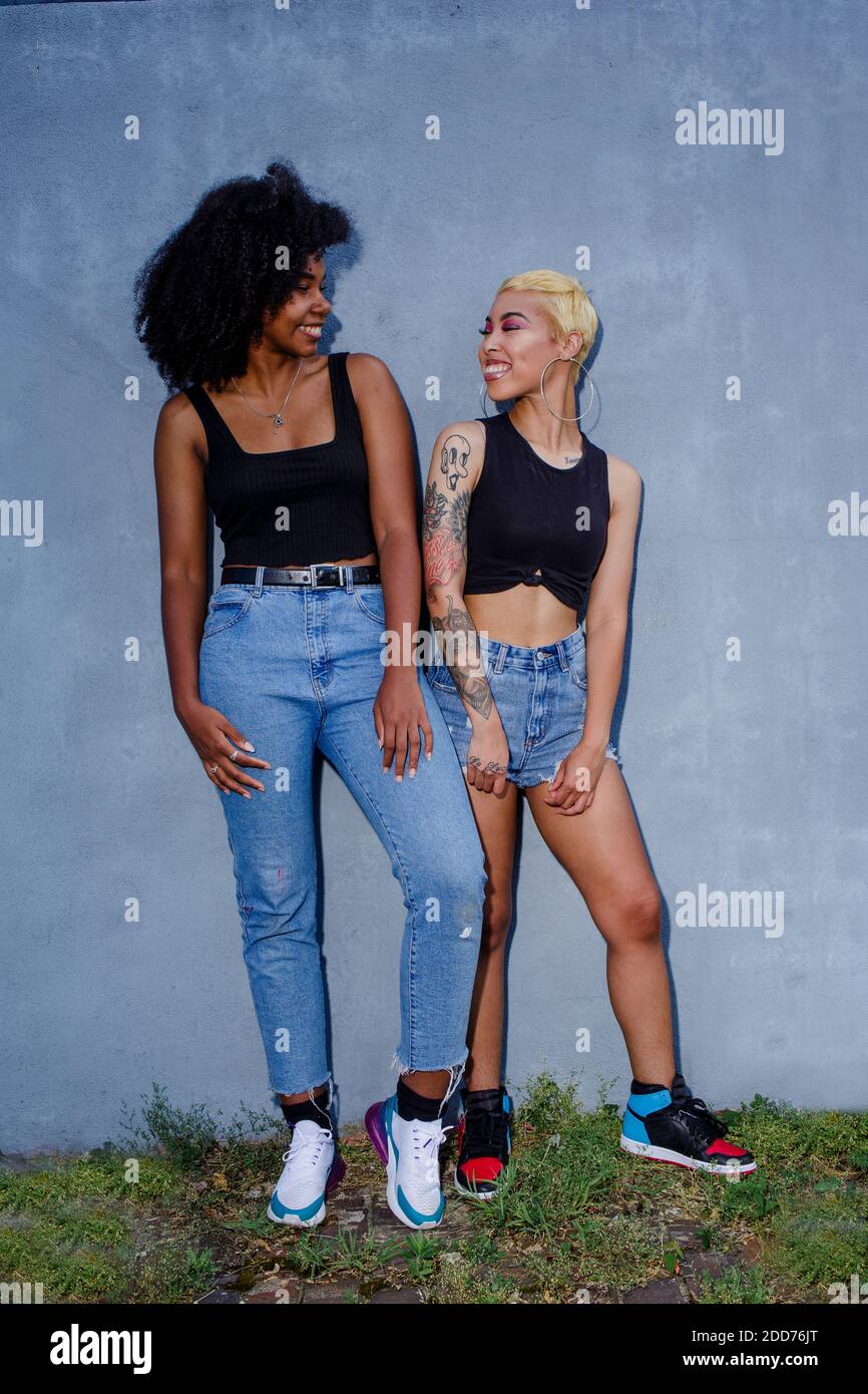 Two smiling friends lean together against a wall in matching clothes ...