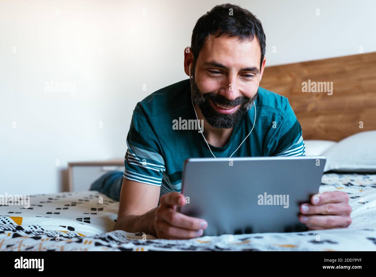 Young man lying on bed looking at digital tablet wearing headphones ...