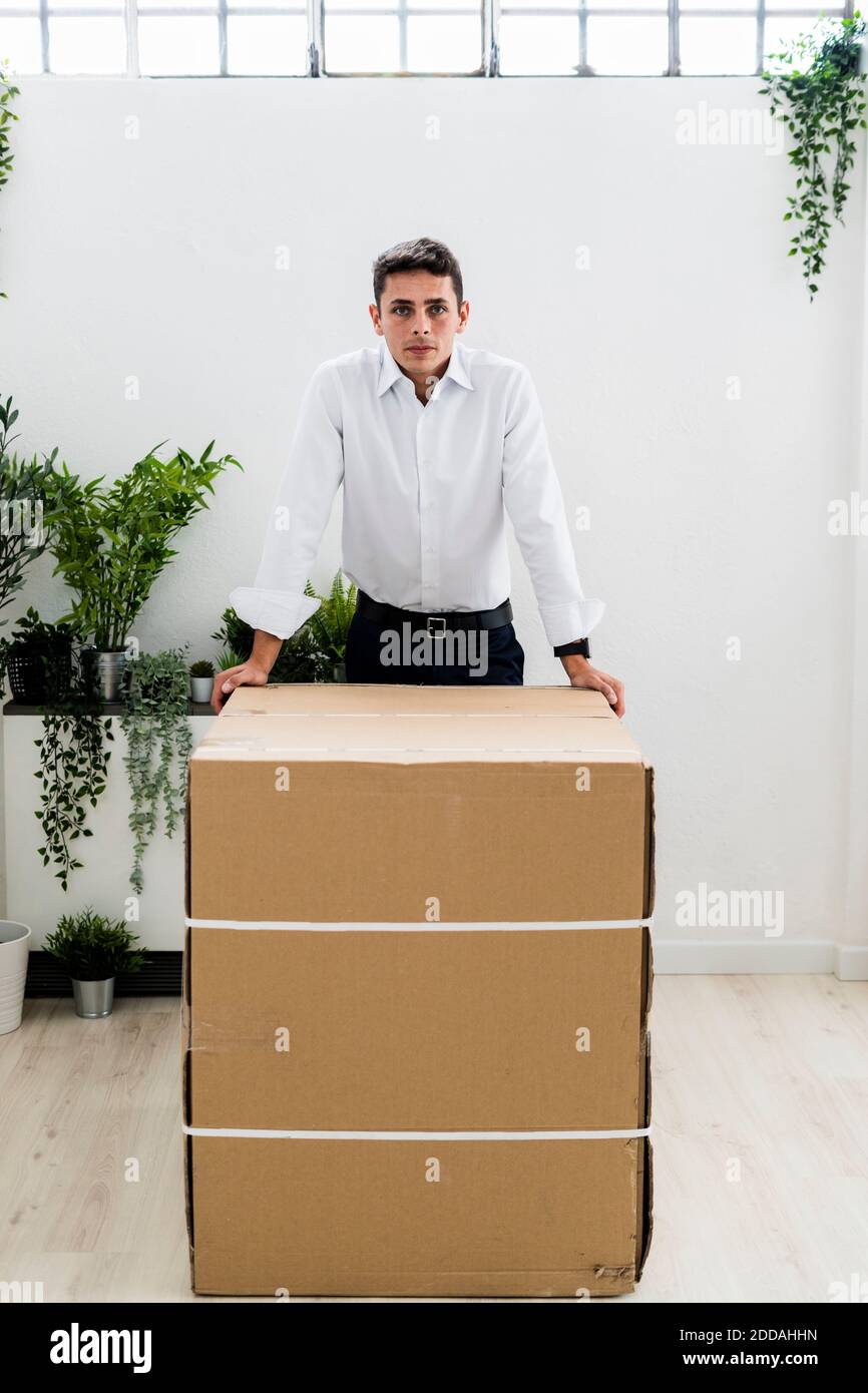 Confident male professional leaning on cardboard box against white wall ...