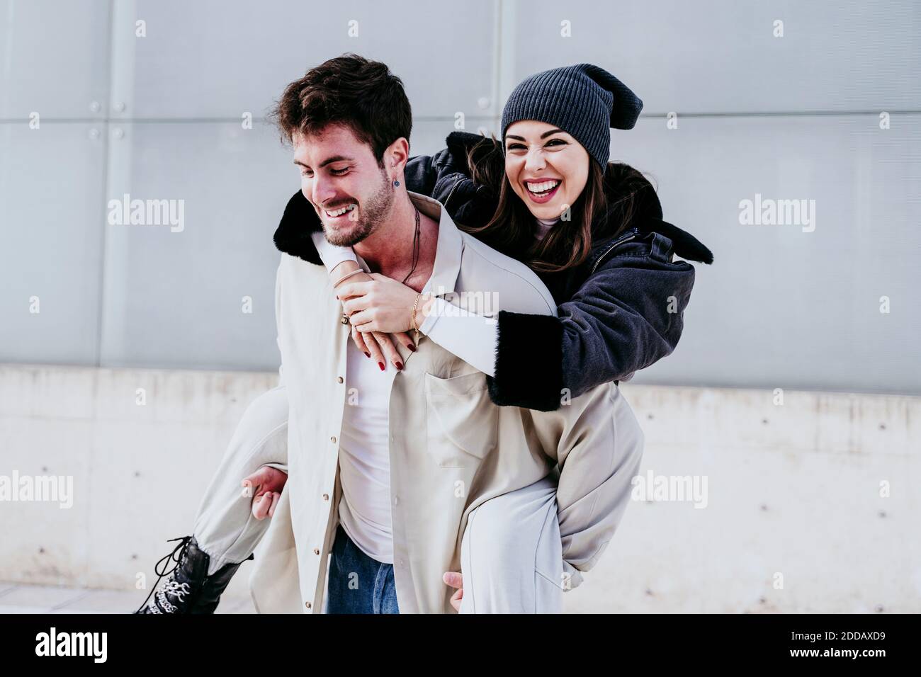 Smiling man piggybacking female partner while standing against wall ...