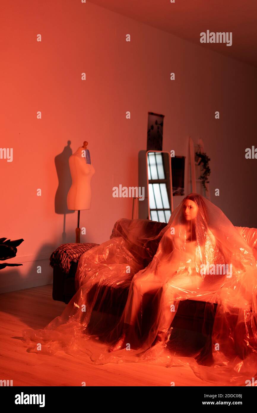 Worried young woman covered in plastic sitting on sofa at home during ...