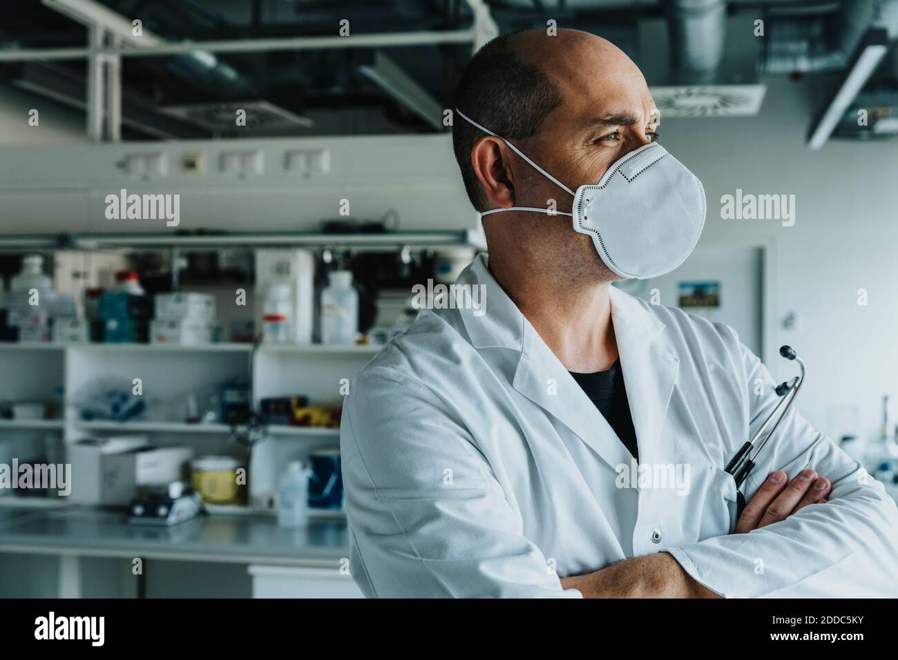 Male scientist wearing face mask looking away while standing with arms ...