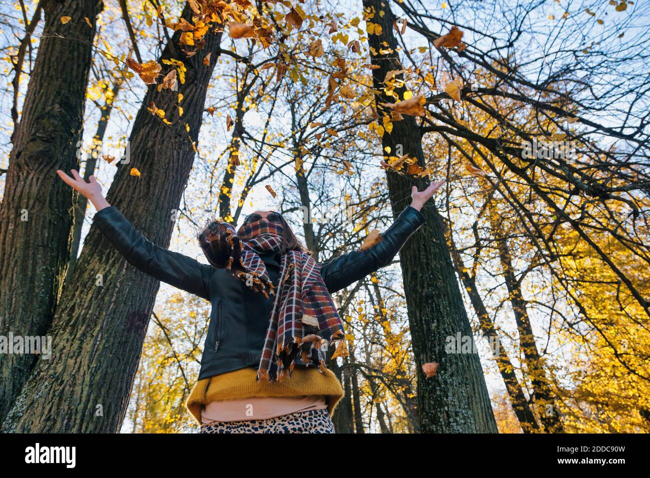 Woman throwing dry autumn leaves while standing with arms outstretched ...