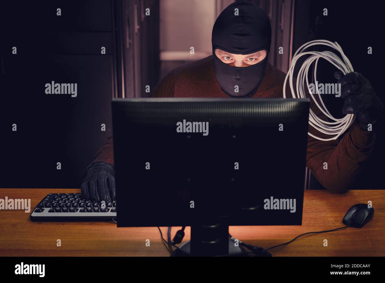 A man in a black mask with wires in his hand sits at a computer in a ...