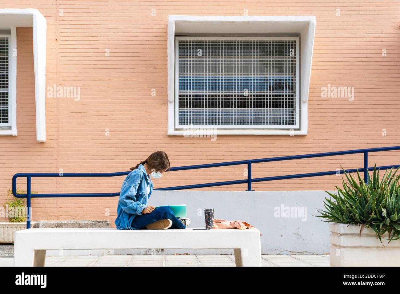 Teenage girl wearing protective face mask reading book while sitting on ...