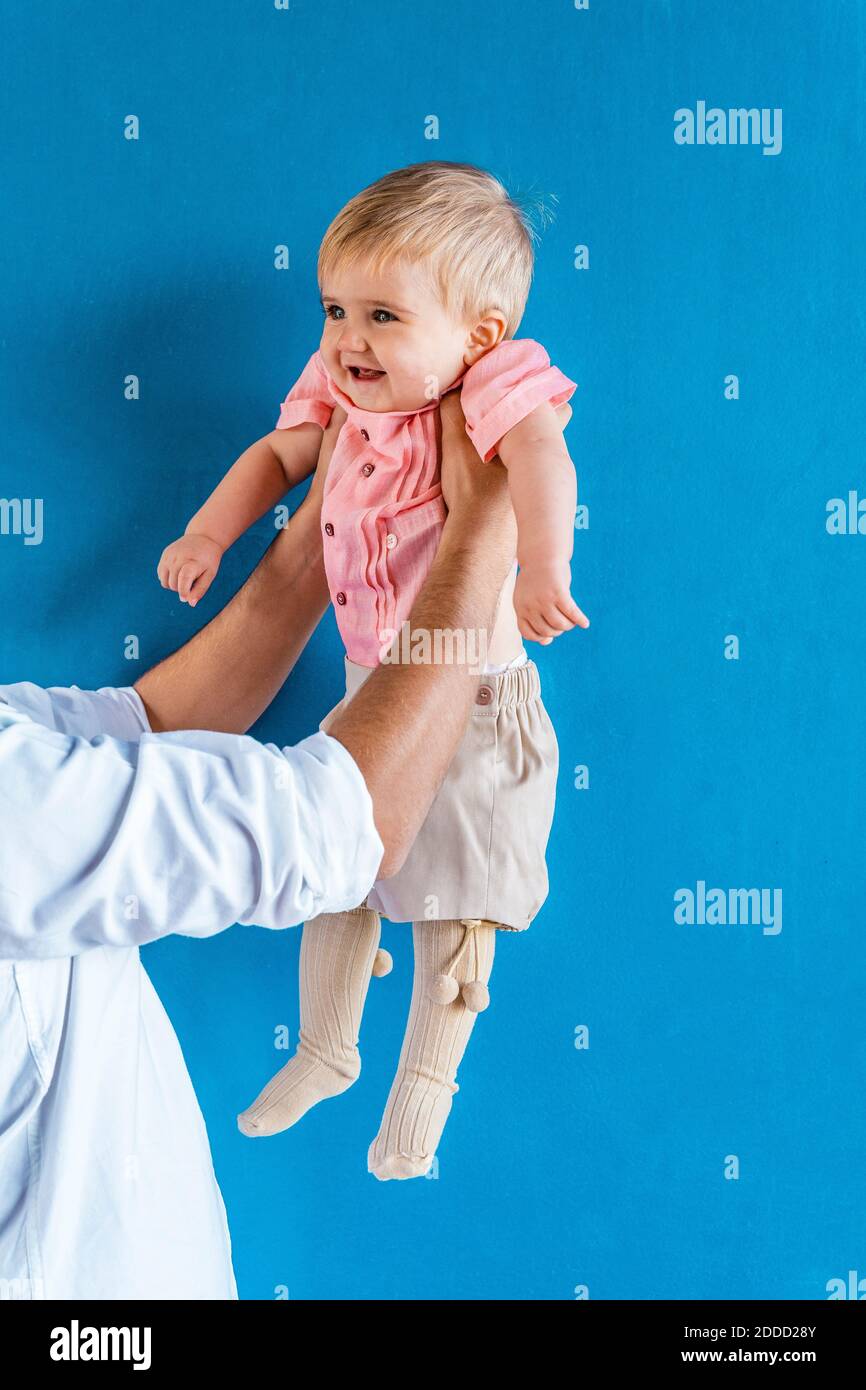 Father picking up baby boy while standing against blue wall Stock Photo ...