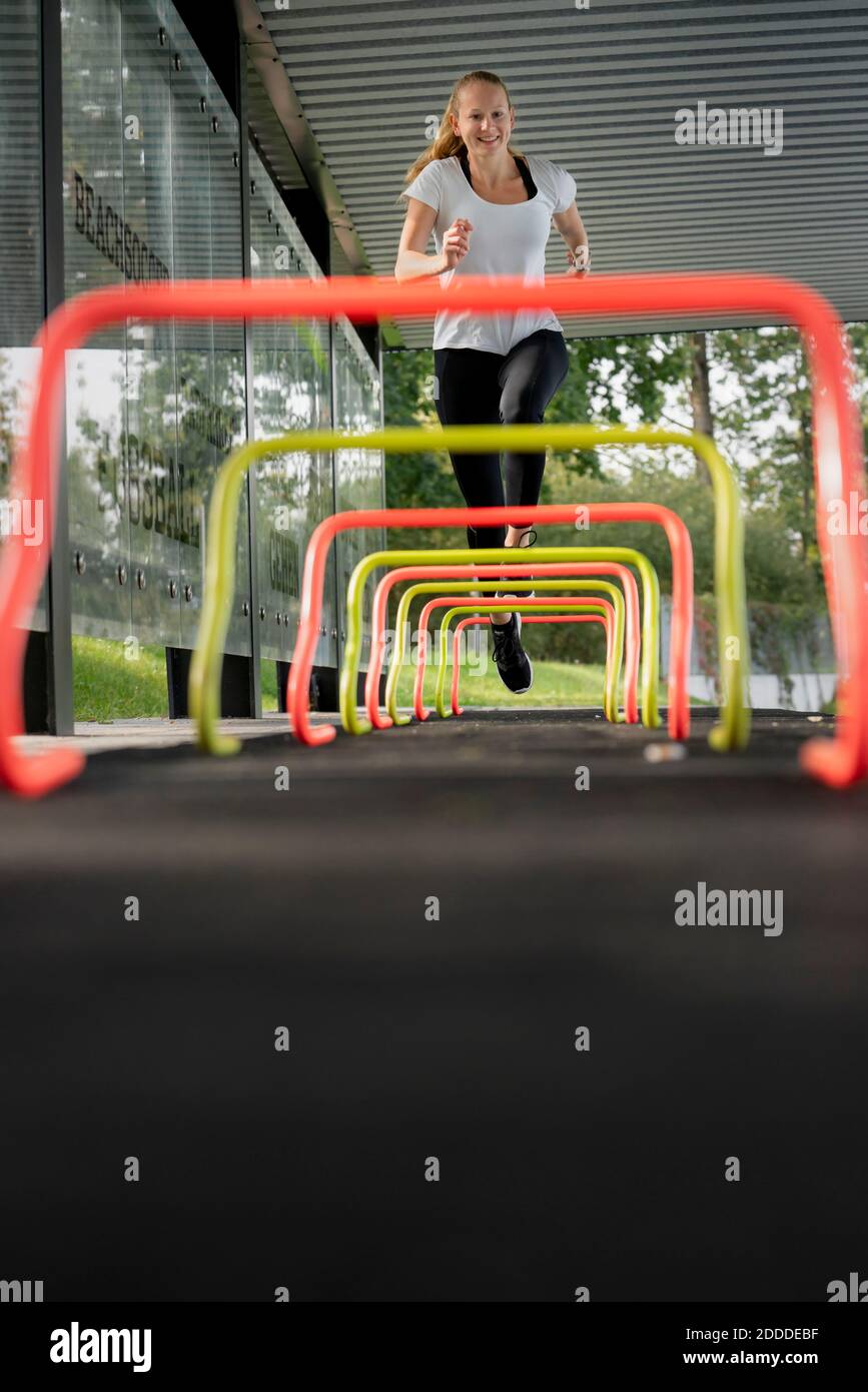 Smiling athlete practicing while running over small hurdle by training ...