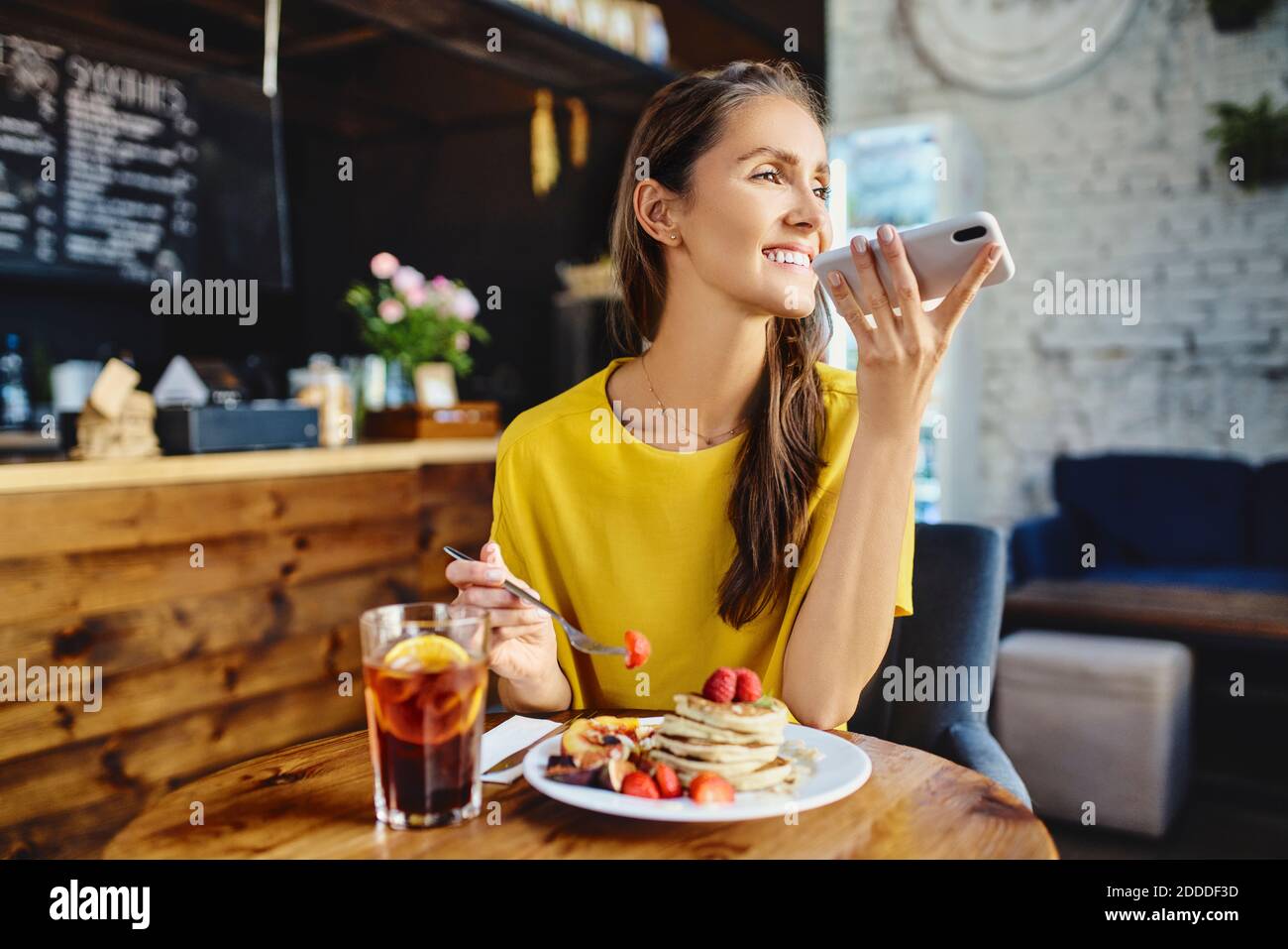 Smiling young female eating berry while using smart phone at table in ...