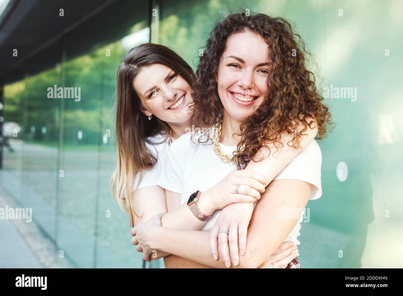 Smiling beautiful woman hugging female friend from behind against wall ...