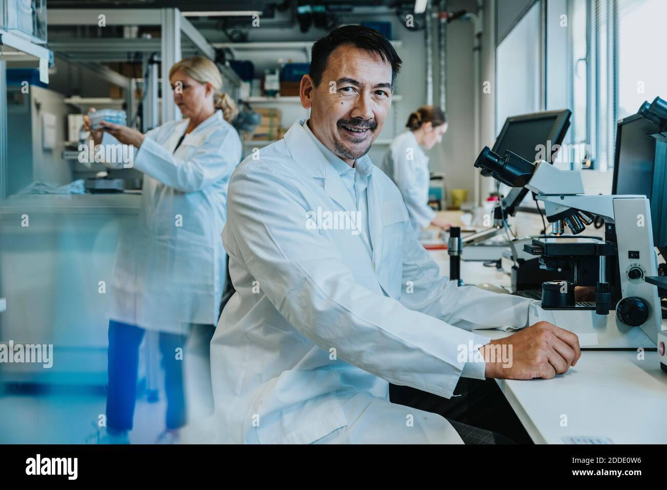 Smiling male scientist sitting by microscope while coworker working in ...